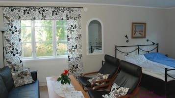 Quarto com cama, escrivaninha com papéis, duas cadeiras, janela com cortinas estampadas e um quadro na parede.