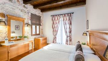 Quarto com parede de pedra, vigas de madeira no teto, cama com roupa de cama branca, cómoda de madeira e um espelho.