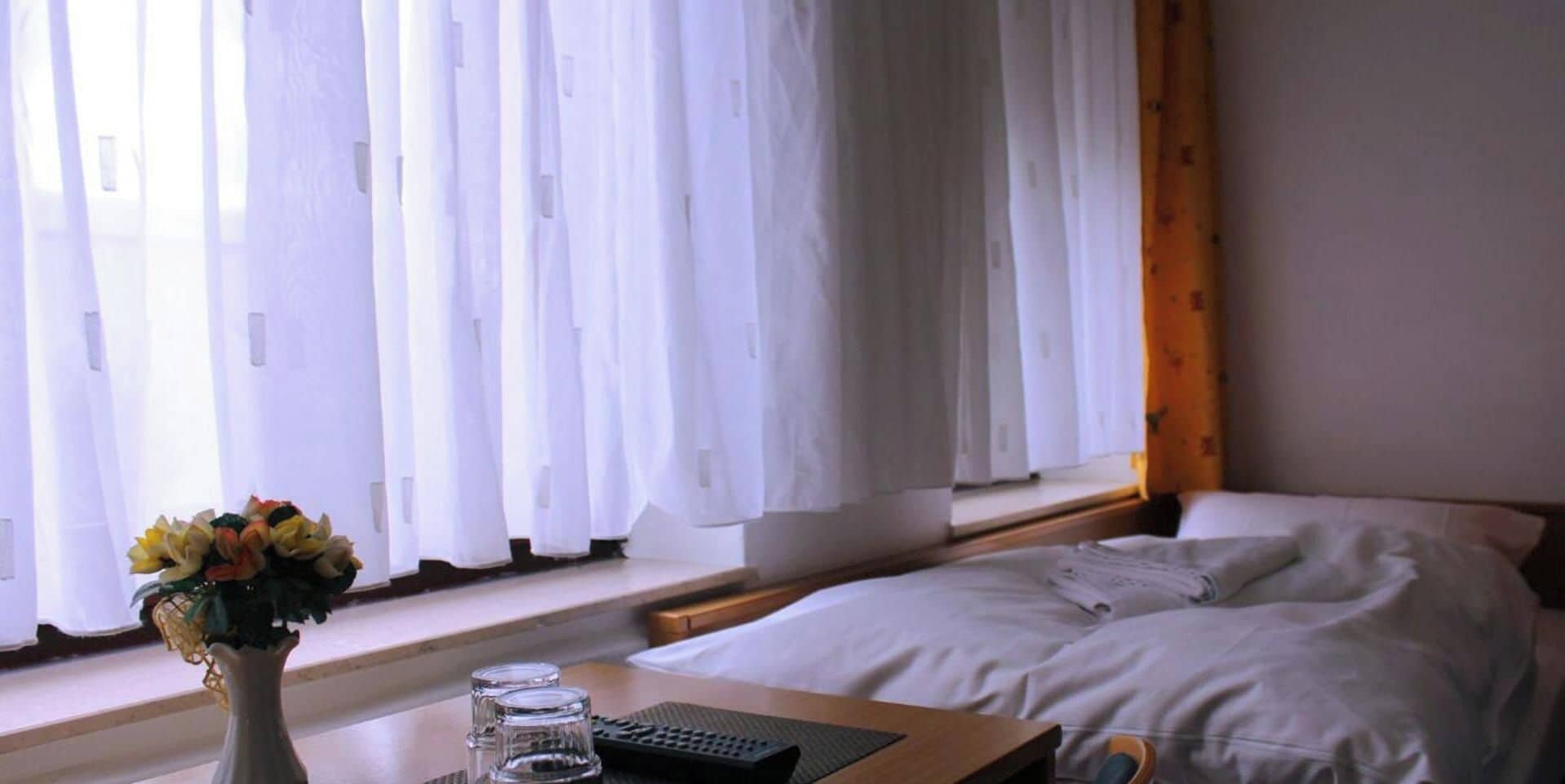 Quarto de hotel com cama, uma mesinha com um vaso de flores e uma janela com cortinas.