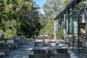 Um pátio externo com móveis de vime e paredes de vidro com vista para um jardim.
