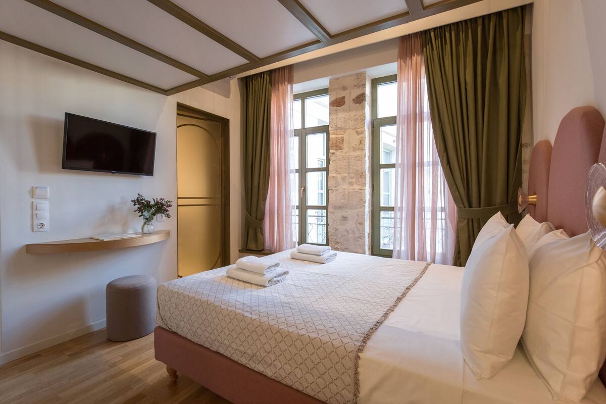 Quarto de hotel com cama, televisão de tela plana na parede, uma pequena planta em uma prateleira e uma janela com cortinas.