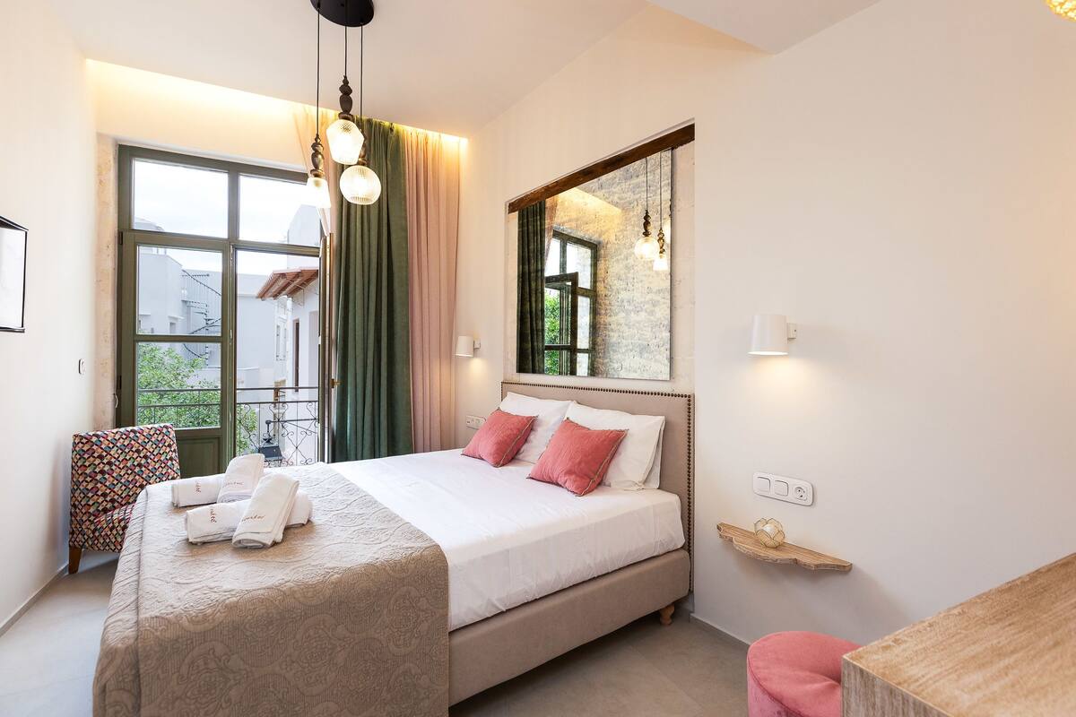 Quarto com uma cama grande, mesa de cabeceira com toalhas, uma cadeira, mesa lateral de madeira e um espelho na parede.