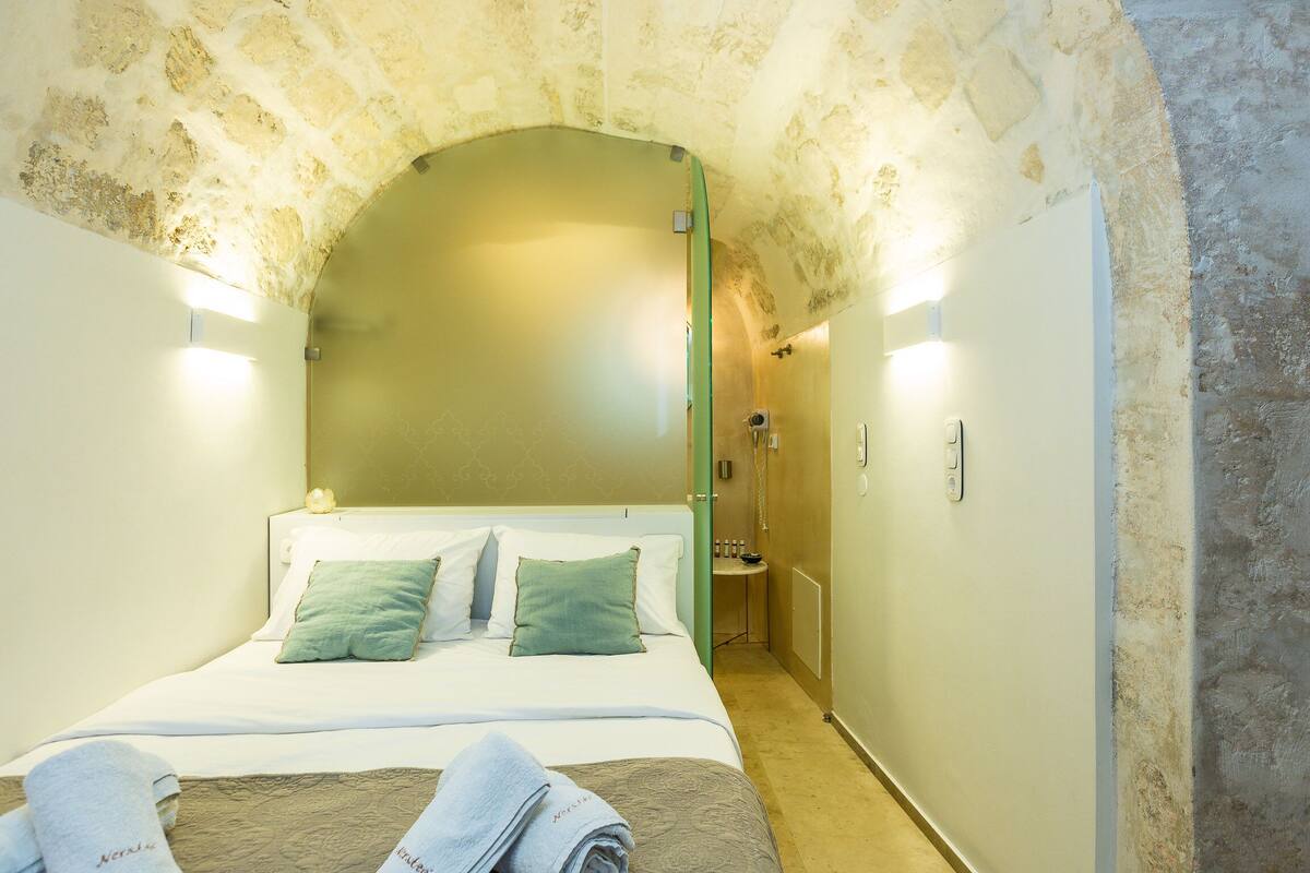 Quarto com teto em arco de pedra, cama com almofadas verdes, mesa de cabeceira pequena com abajur e uma toalha na cama.
