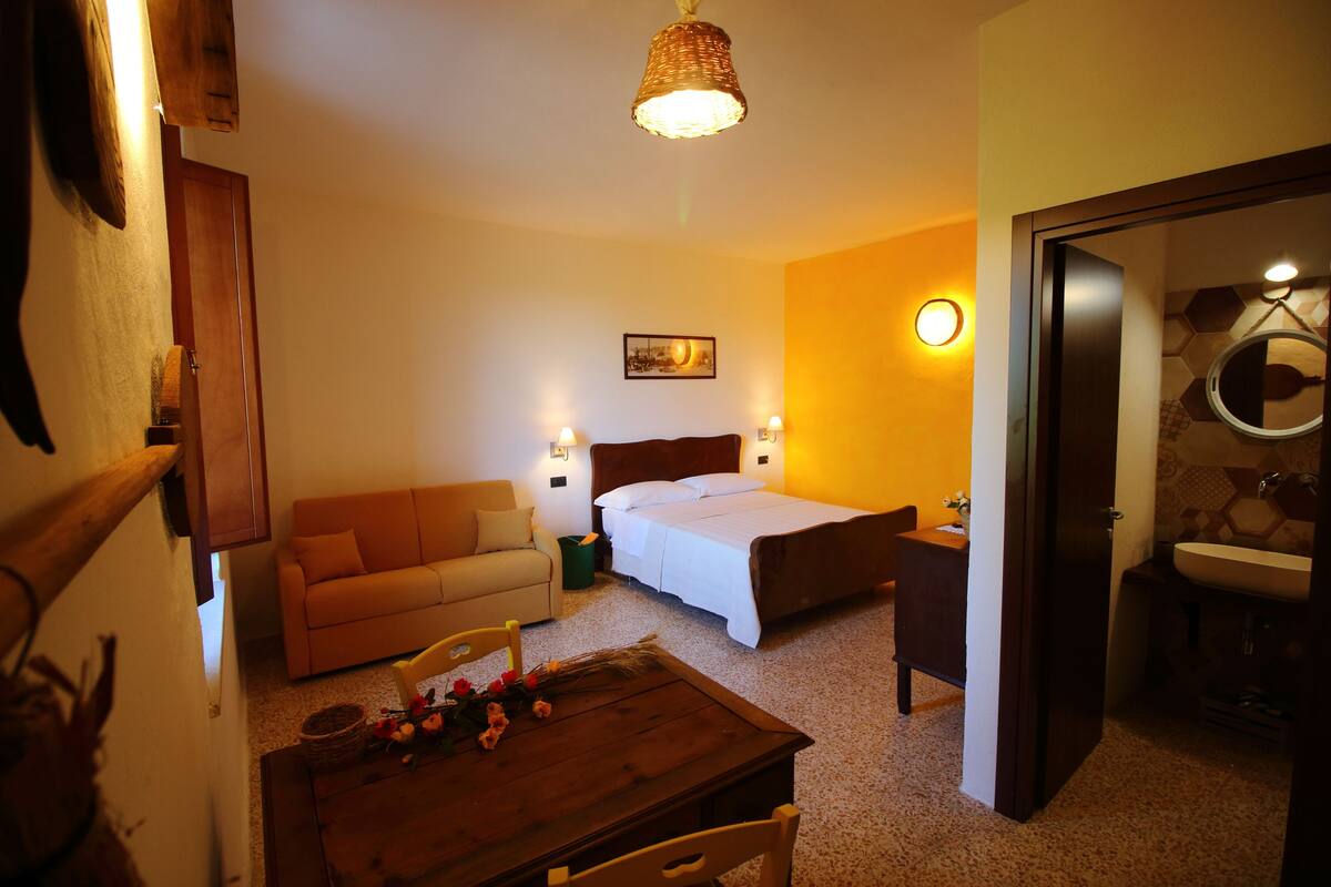 Quarto com cama, sofá, mesa com flores, mesa de cabeceira, abajur e banheiro com pia e espelho.