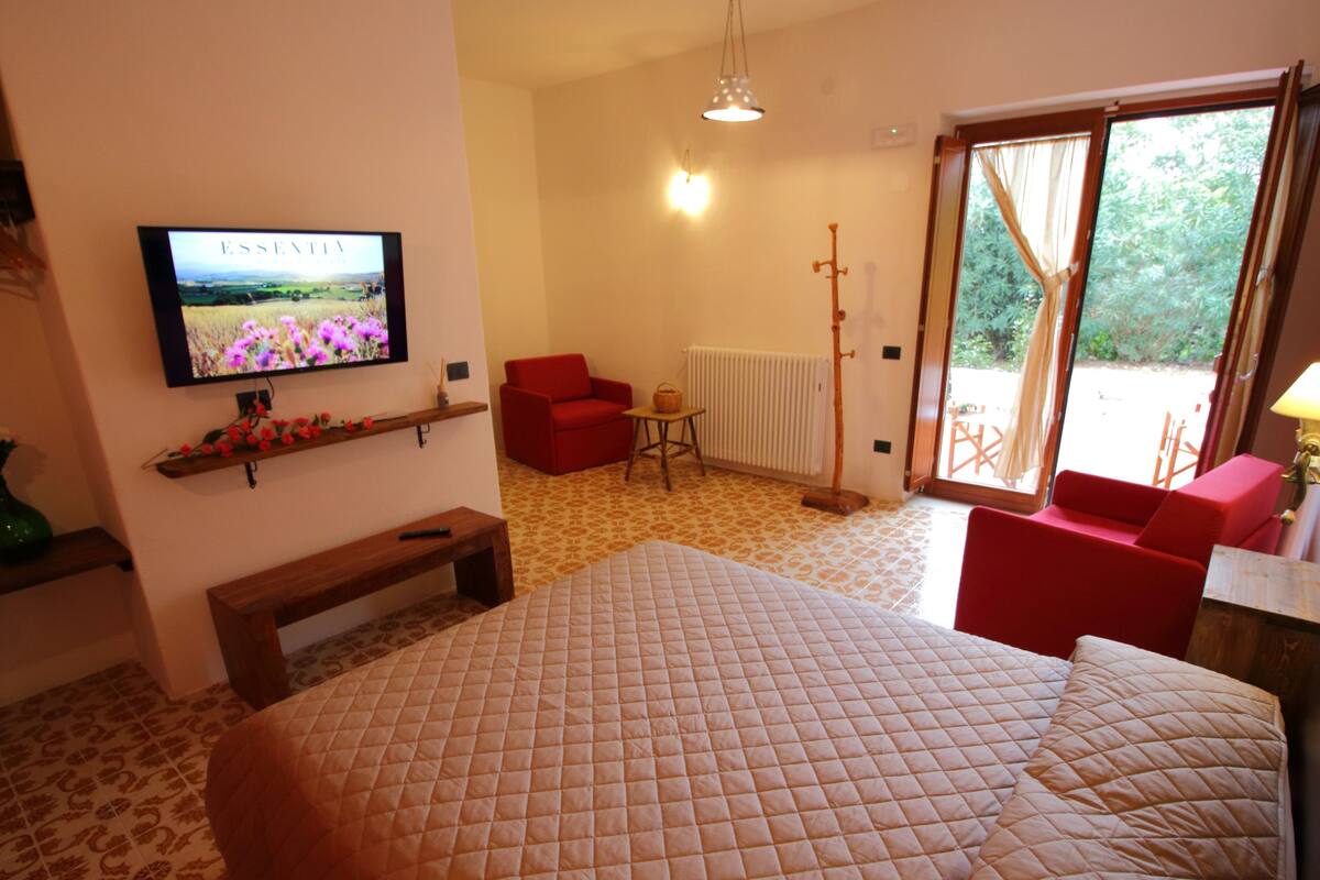 Quarto com cama, TV na parede, uma cadeira vermelha, uma mesa pequena e vista através de uma porta de vidro.