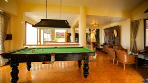 Uma mesa de bilhar em um ambiente com piso de padrão hexagonal, poltronas de couro e uma grande janela que oferece luz natural.