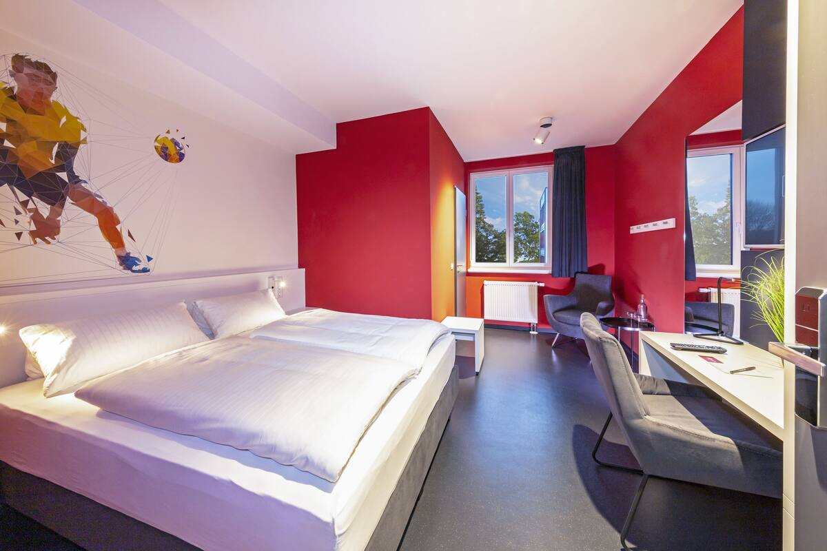 Quarto de hotel moderno com cama, escrivaninha e cadeira, uma janela grande e uma obra de arte com tema esportivo.