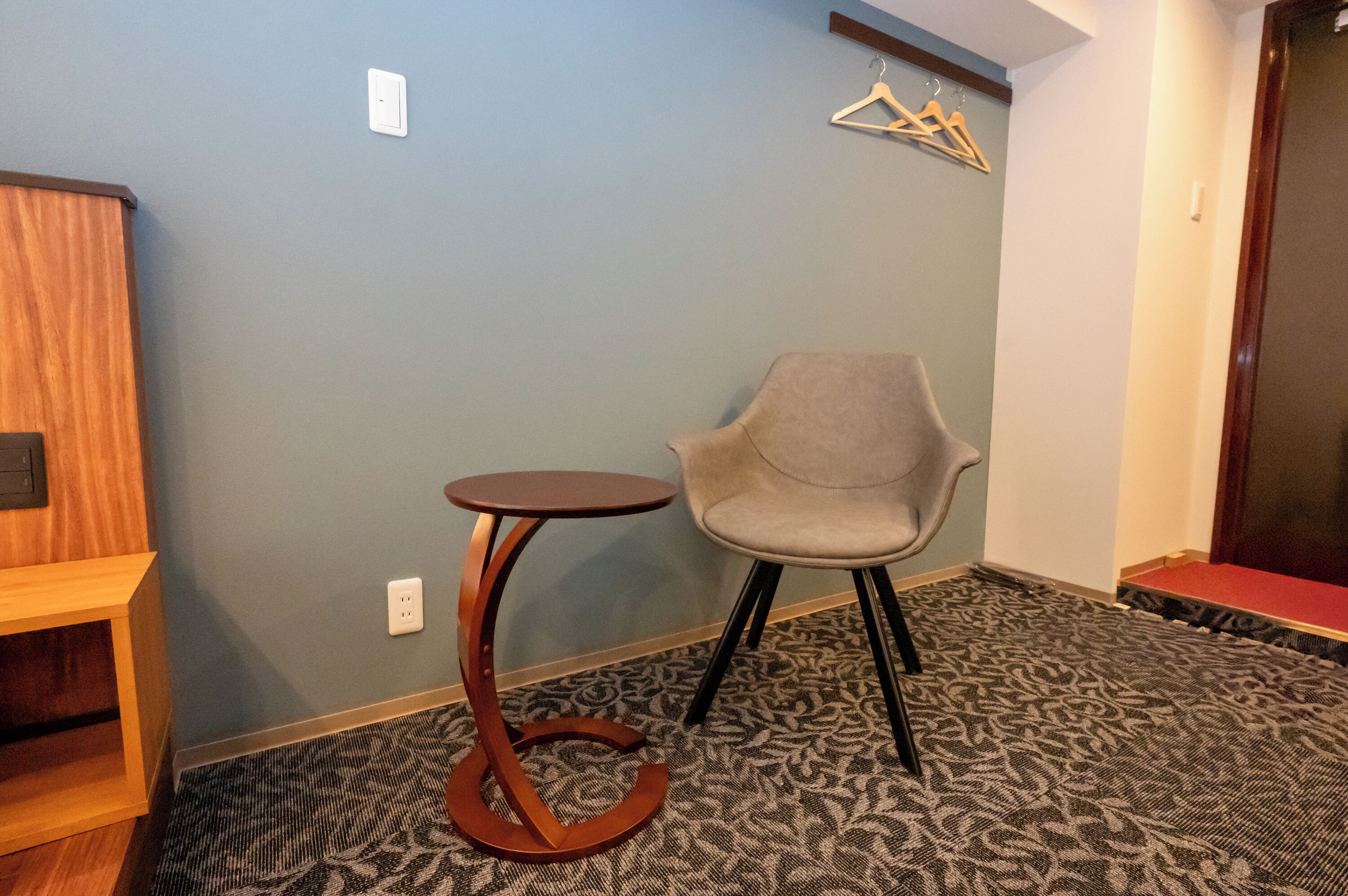 Um quarto com carpete estampado, uma mesa redonda com base curva, uma cadeira estofada cinza e um armário de madeira.