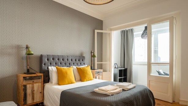 Quarto bem arrumado com cabeceira estofada, mesa de cabeceira de madeira, cama com colcha cinza e almofadas amarelas, abajur e janela com cortinas.