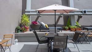 Pátio externo com mesa e cadeiras, cercado por plantas em vasos e um grande guarda-sol.