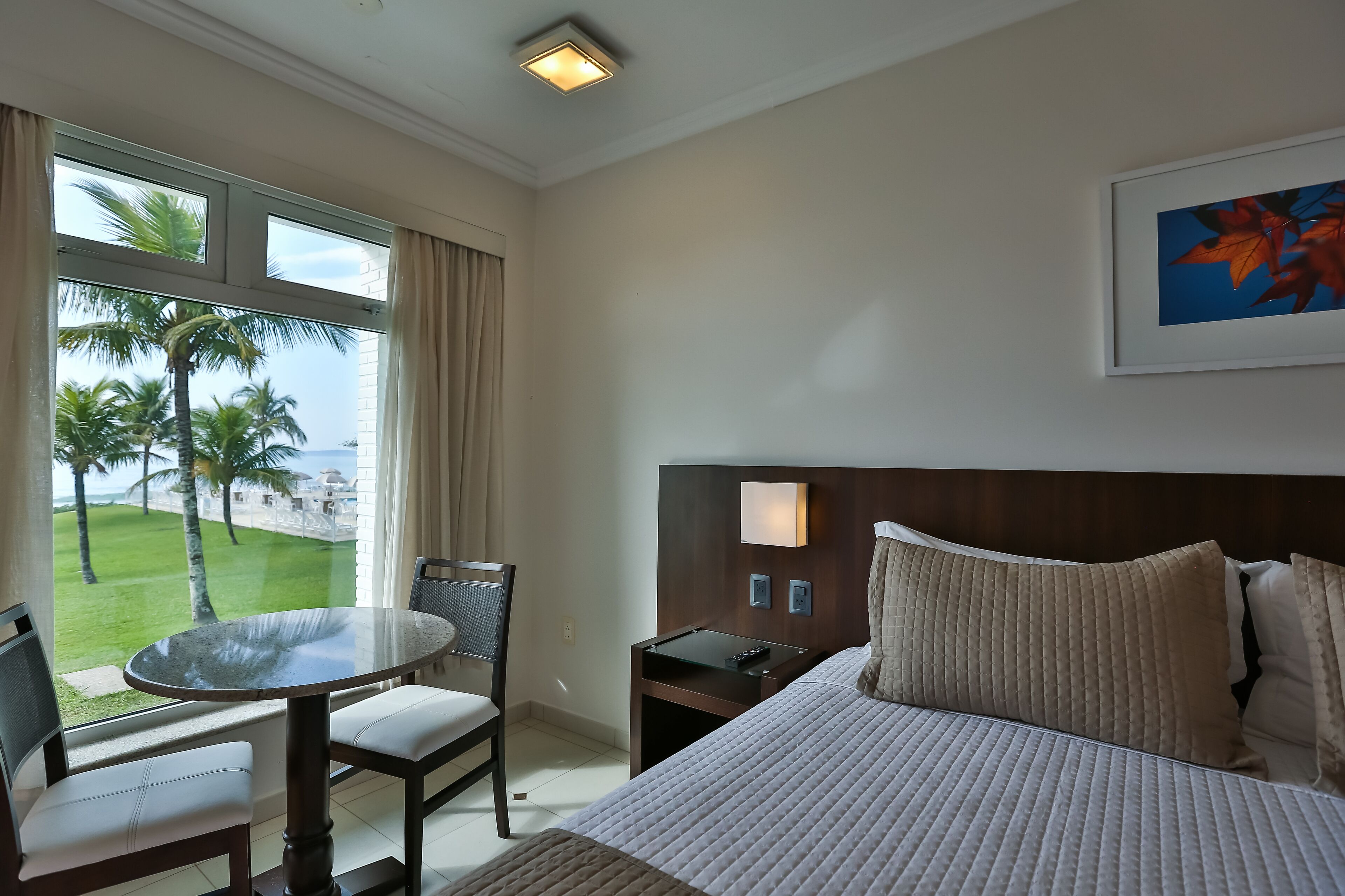 Quarto de hotel com cama, uma pequena mesa, cadeira, mesa de cabeceira, janela com vista para palmeiras e uma praia, e um quadro na parede.