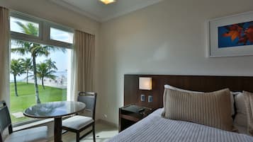 Quarto de hotel com cama, uma pequena mesa, cadeira, mesa de cabeceira, janela com vista para palmeiras e uma praia, e um quadro na parede.