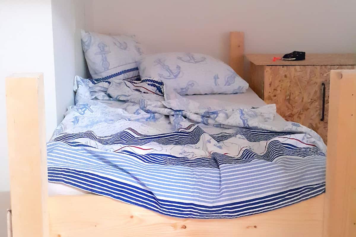 Quarto pequeno com uma cama de solteiro, estrutura de madeira, piso de azulejos brancos e uma mesinha de cabeceira simples com abajur.