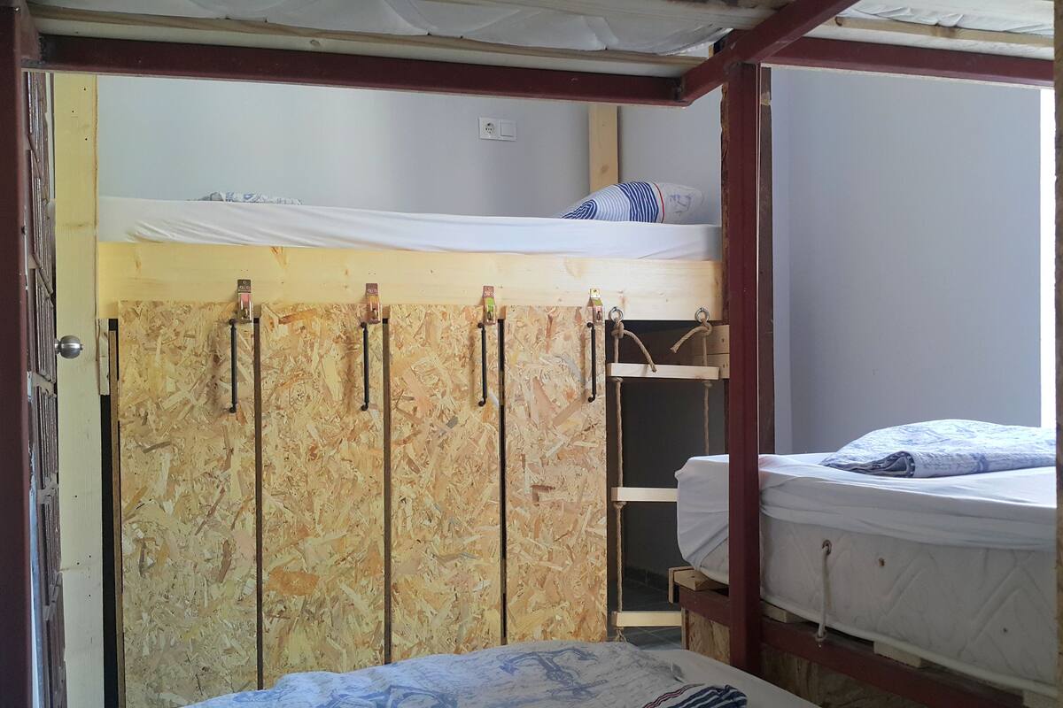 Quarto pequeno e compacto com uma cama de solteiro e uma cama individual. Há uma porta de madeira com puxadores de metal e um pequeno estante acima dela.