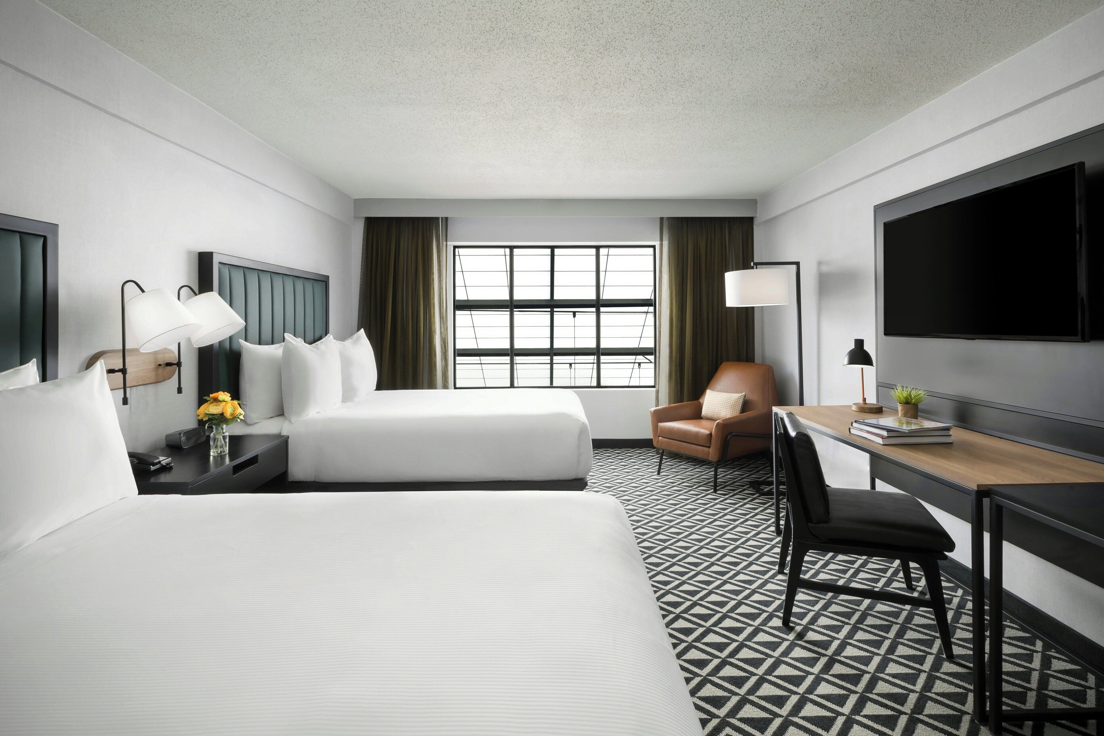 Quarto de hotel com duas camas, uma mesa, uma cadeira, uma televisão e uma janela com cortinas.