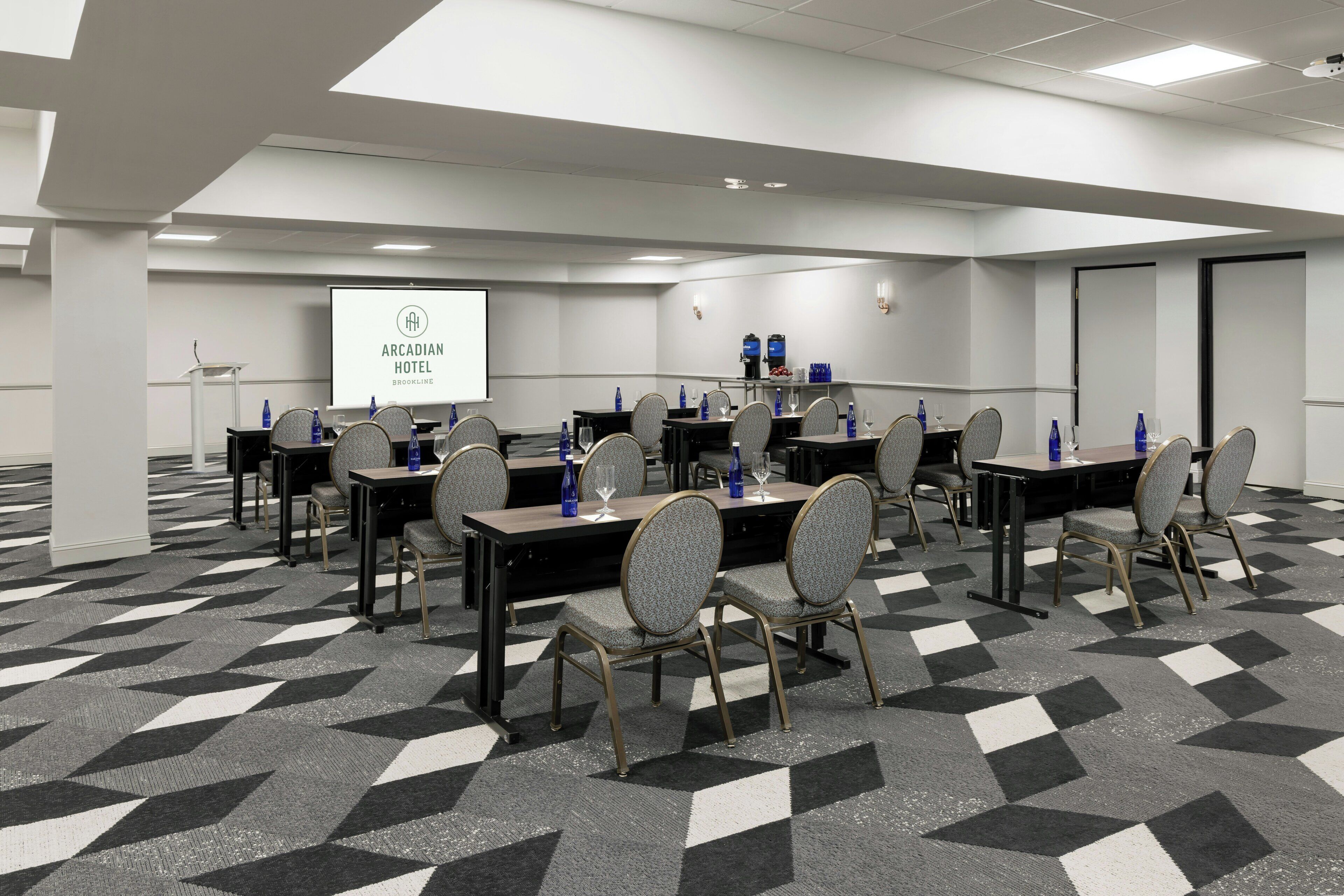 Uma sala de conferências com tela de projetor, mesas, cadeiras e garrafas de água.