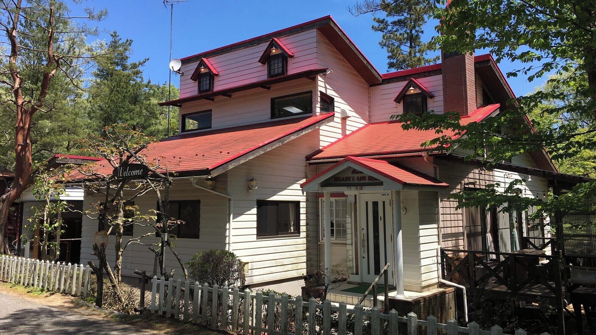 Uma casa de dois andares com telhado vermelho, cerca branca de piquete e um letreiro com a palavra 'Welcome'.