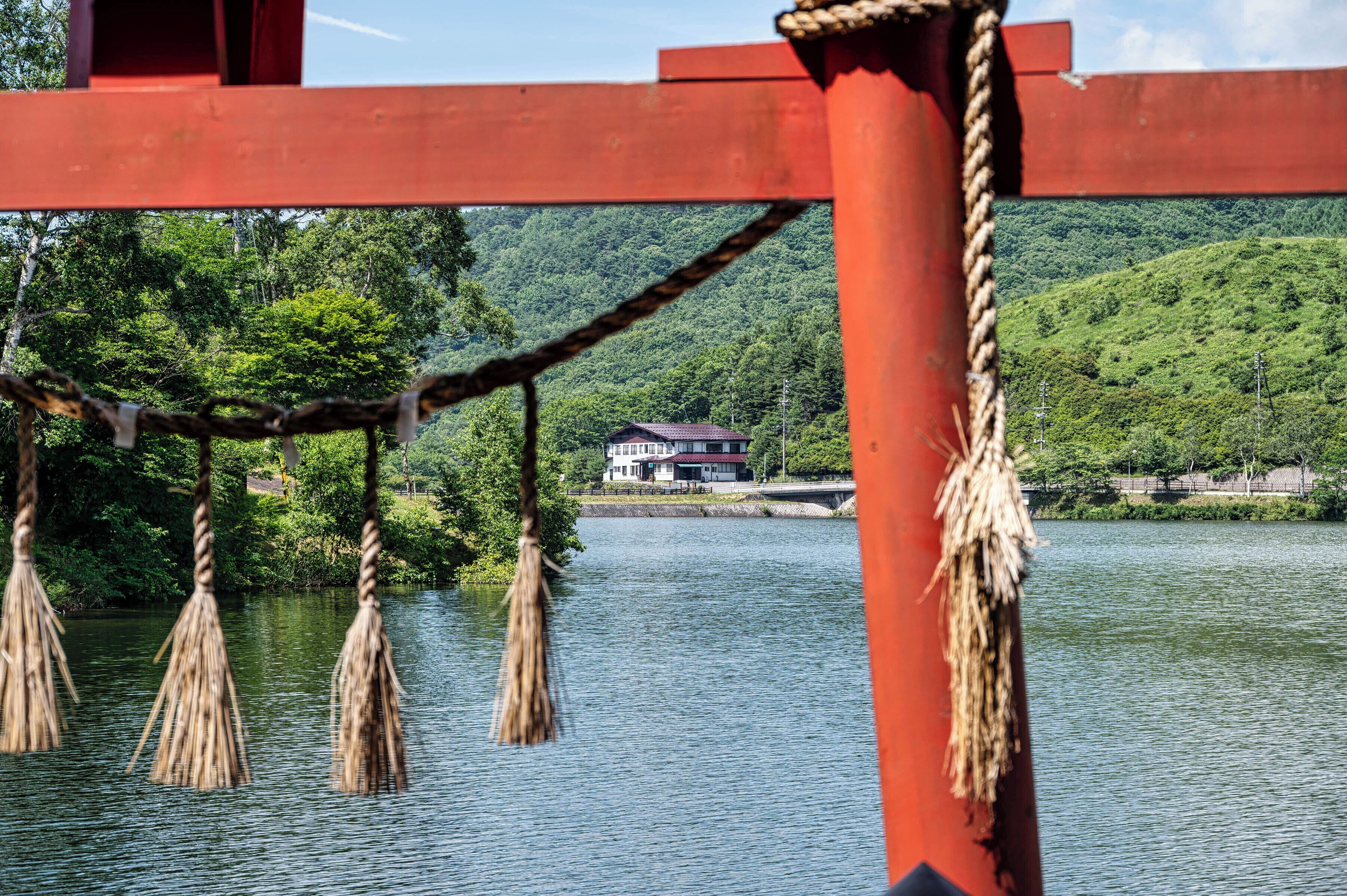 Um tradicional portão torii japonês com corda e decorações de palha, com vista para um rio calmo, vegetação exuberante e uma casa ao fundo.