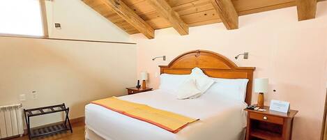 Quarto com teto de madeira, uma cama grande com roupa de cama branca, duas mesas de cabeceira com abajures e um radiador.
