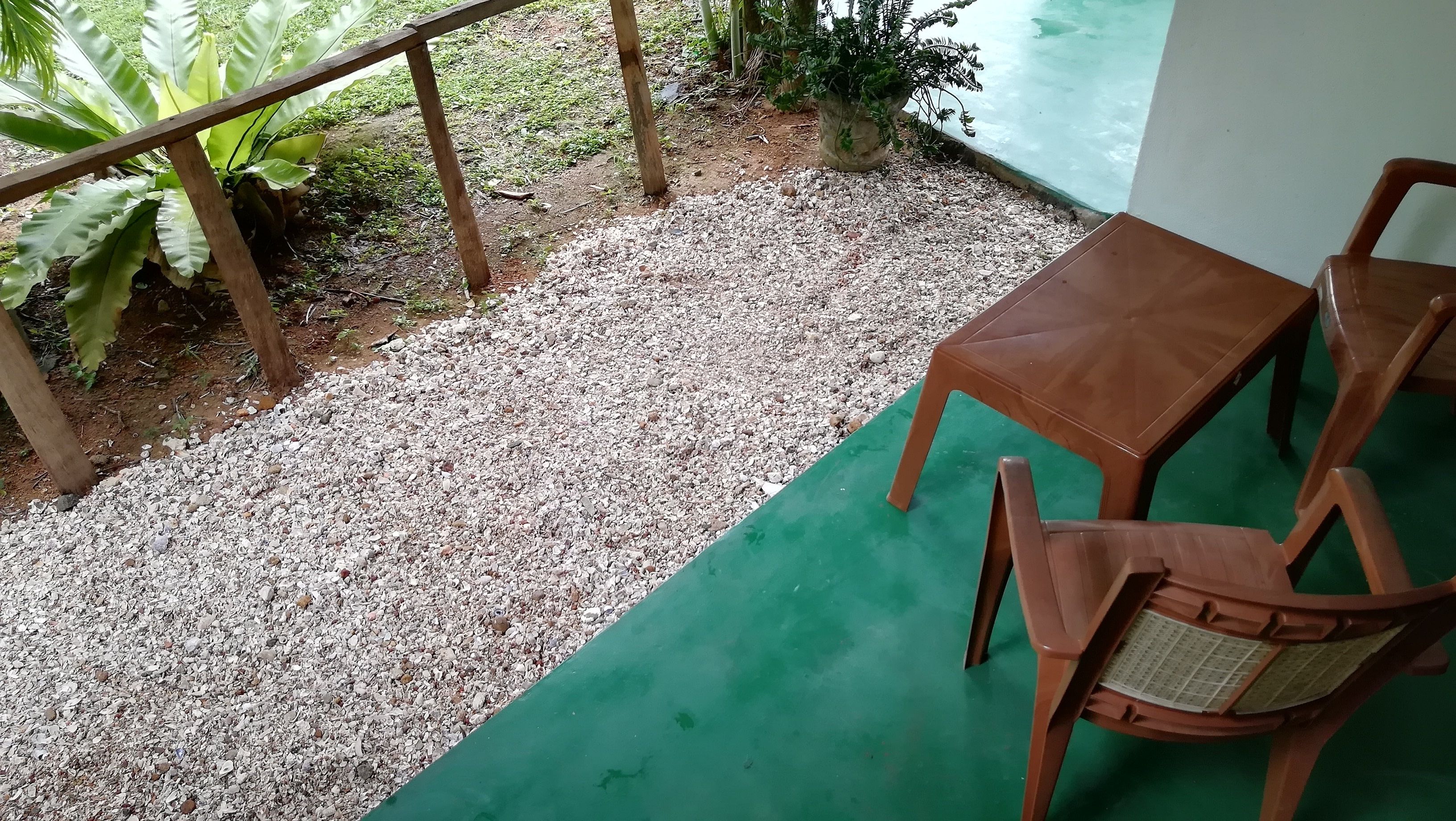 Pátio com mesa e cadeira de madeira, rodeado por cascalho e plantas.