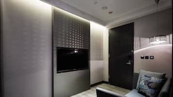Quarto de hotel moderno com televisão de tela plana na parede, porta escura, piso de madeira claro e sofá escuro com almofada estampada.