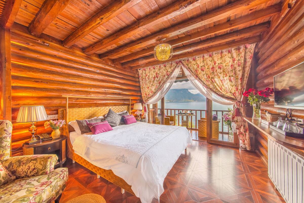 Quarto de cabana de madeira com uma cama grande, uma mesa de cabeceira, uma cadeira e vista para as montanhas pela janela.