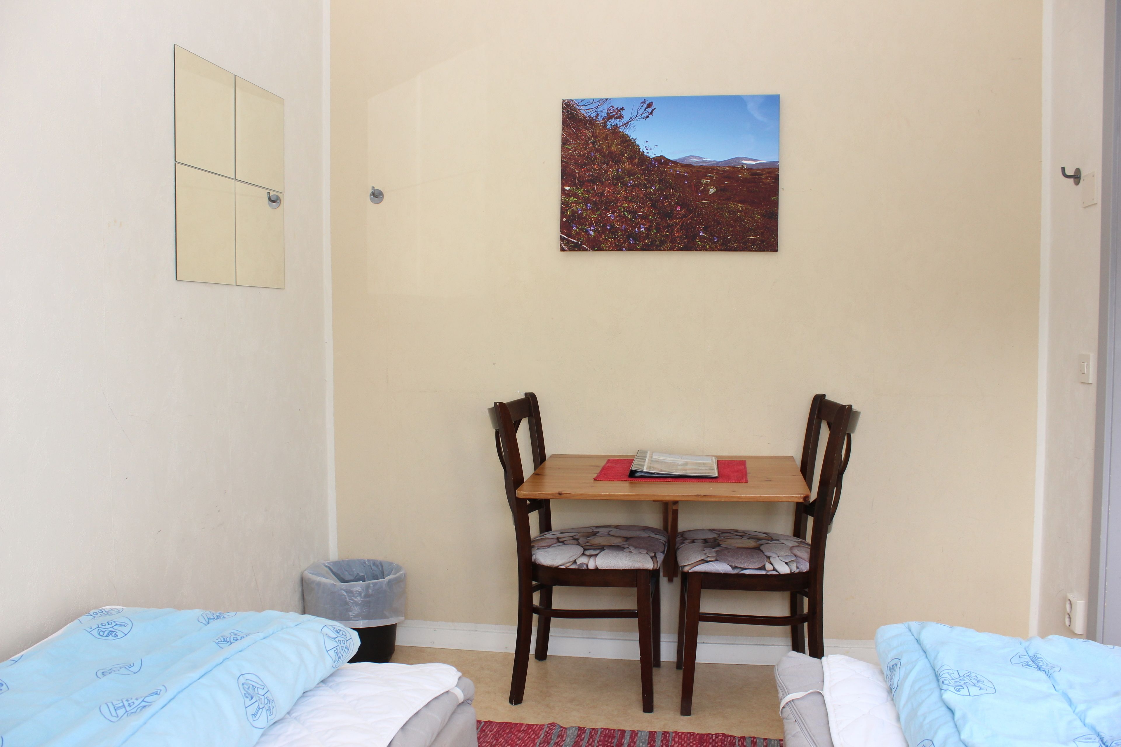 Quarto com duas camas, uma mesa de madeira com cadeiras, um quadro na parede e uma lixeira.