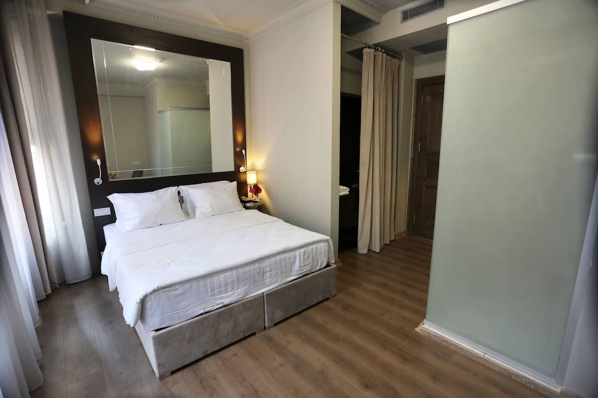 Quarto de hotel com uma cama grande, um espelho, mesa de cabeceira com abajur e piso de madeira.