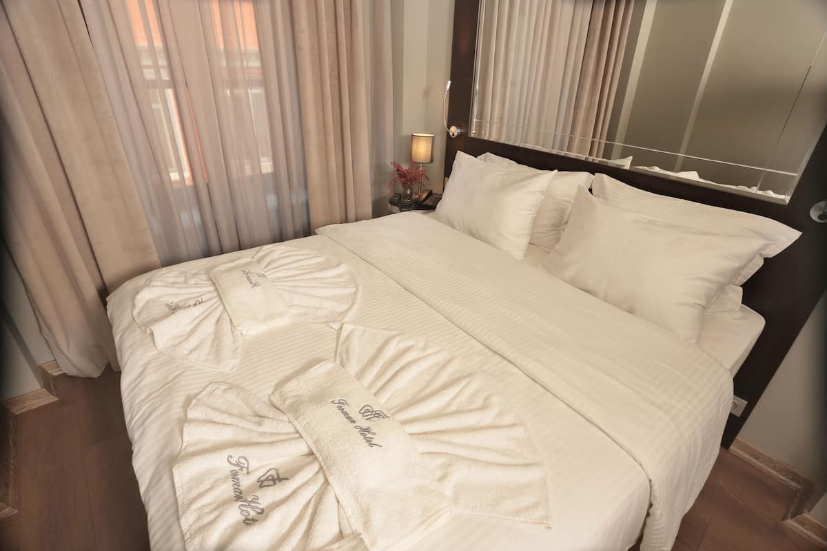 Uma cama bem arrumada com edredom branco e almofadas, uma mesa de cabeceira com abajur e cortinas translúcidas.