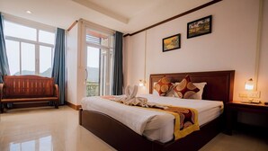 Quarto de hotel com uma cama grande, um banco de madeira, uma varanda com cortinas e duas quadros na parede.