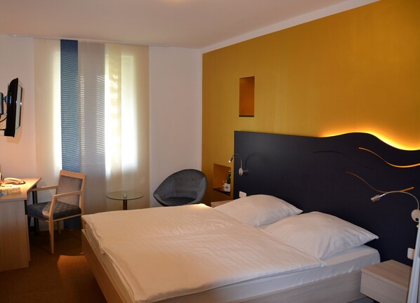 Quarto de hotel com cama, escrivaninha com cadeira, mesa pequena e janela com persianas.