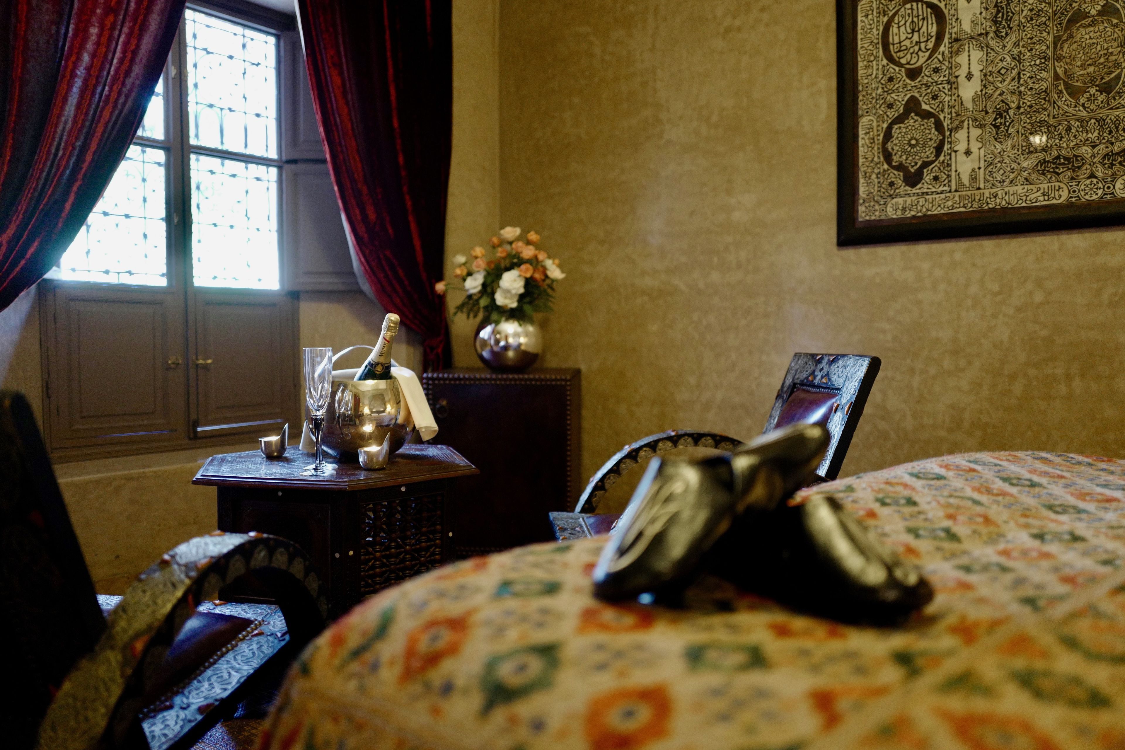 Quarto de hotel com cama, uma cadeira, uma mesinha com garrafa e copos, um vaso com flores e um quadro decorativo na parede.