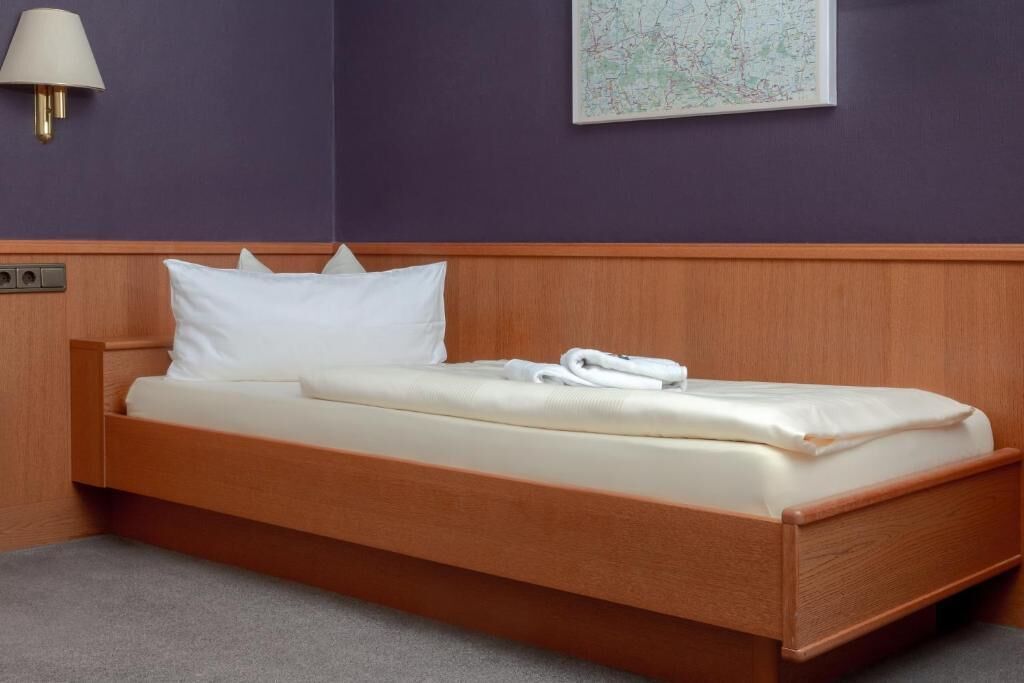 Uma cama de solteiro com roupa de cama branca, uma luminária fixada na parede e um mapa emoldurado.