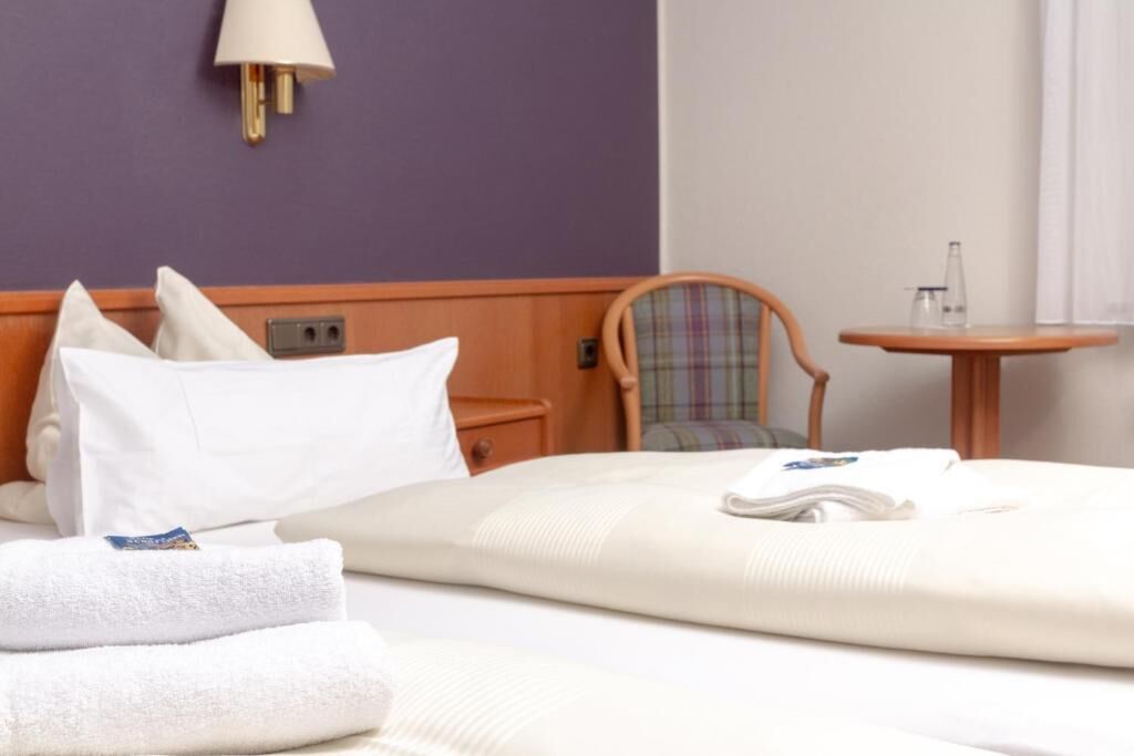 Quarto de hotel com cama, almofadas, poltrona, mesa de cabeceira, luminária e uma pequena mesa com um copo.