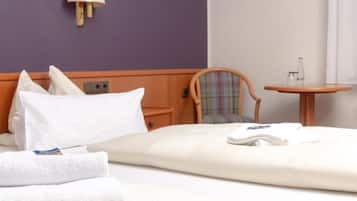 Quarto de hotel com cama, almofadas, poltrona, mesa de cabeceira, luminária e uma pequena mesa com um copo.