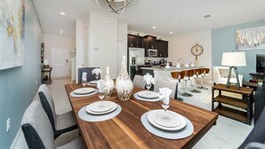 Uma sala de jantar moderna com uma mesa de madeira posta para quatro pessoas, uma cozinha ao fundo e uma sala de estar com sofá e luminária.