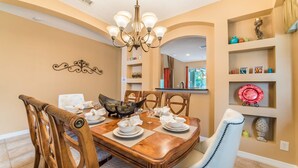 Uma sala de jantar com mesa de madeira posta para seis pessoas, rodeada por cadeiras. Um lustre pendurado no teto e prateleiras com objetos decorativos nas paredes.
