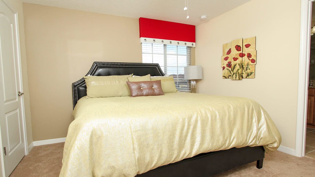 Quarto com cama, janela com persianas, parede de destaque vermelha e um quadro na parede.