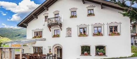 Um edifício branco com varanda, várias janelas com caixas de flores e um espaço de restaurante com mesas ao ar livre.