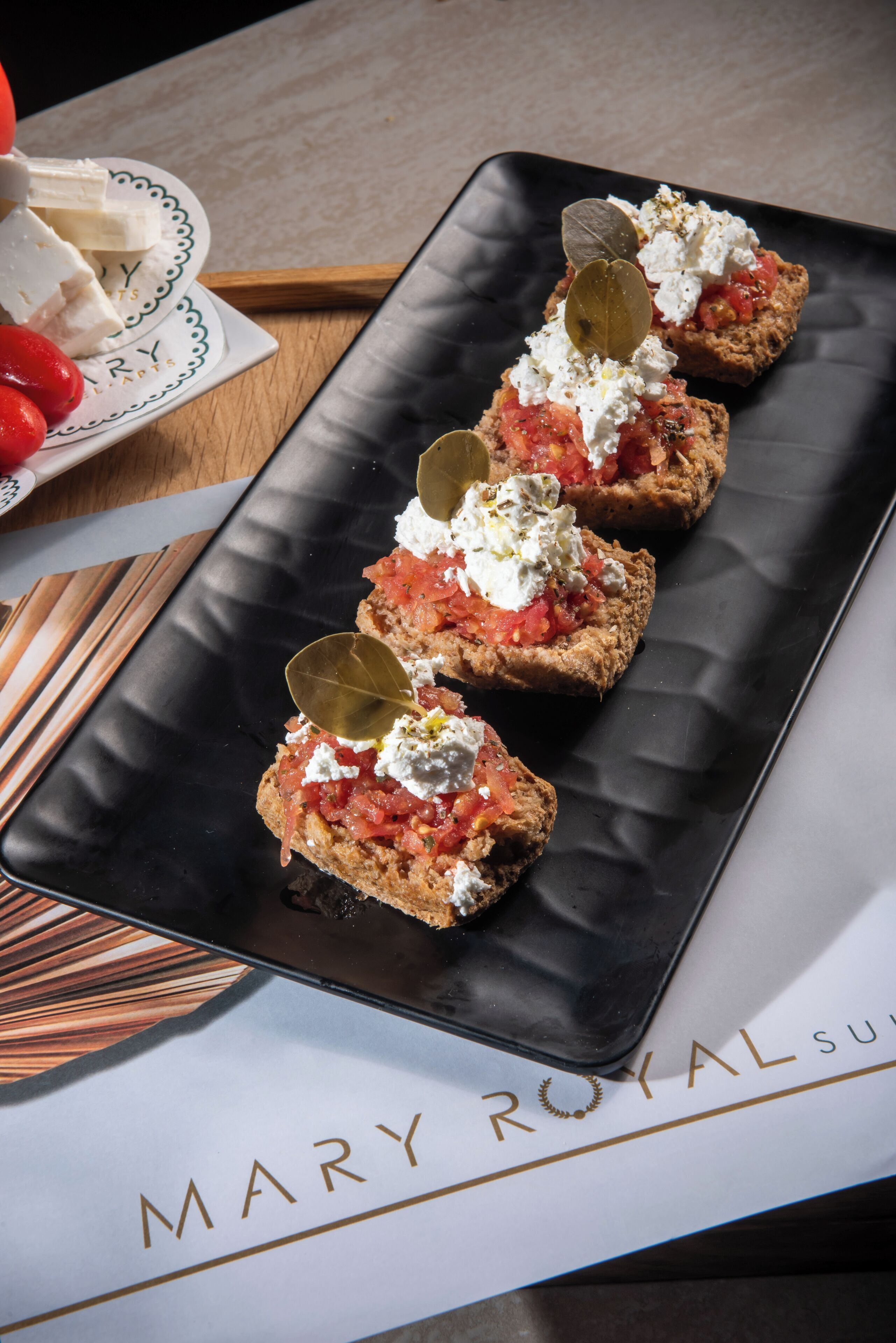 Uma placa preta retangular com três bruschettas cobertas de tomate, queijo e folhas de louro.