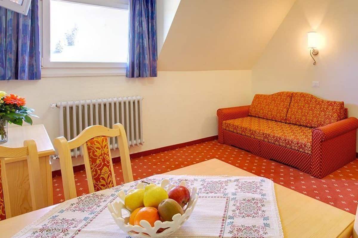 Um quarto acolhedor com uma mesa de jantar posta com frutas, um sofá vermelho estampado e uma cama com cabeceira de madeira.