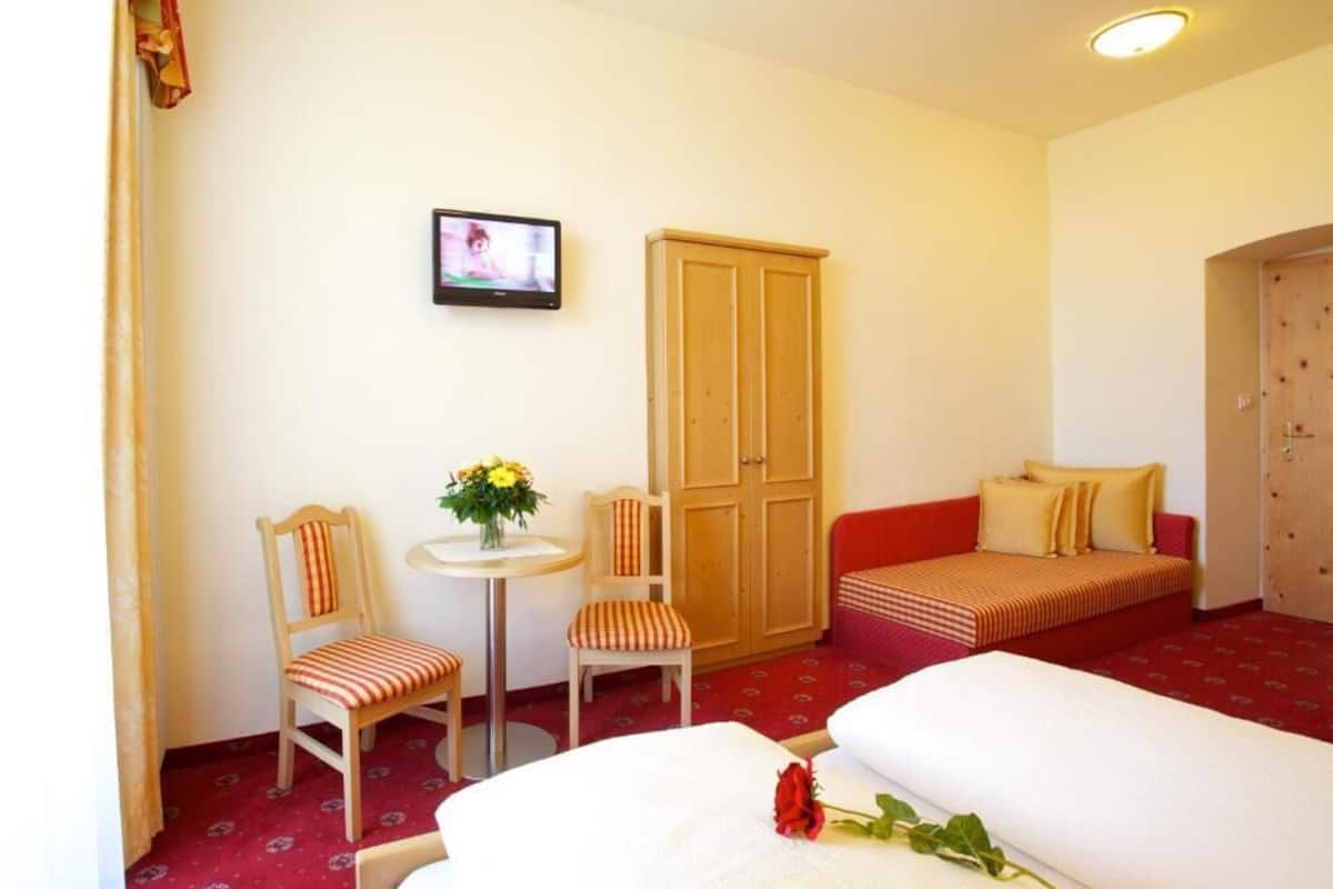 Quarto de hotel com cama, uma pequena mesa com um vaso de flores, duas cadeiras, um guarda-roupa de madeira e uma televisão na parede.