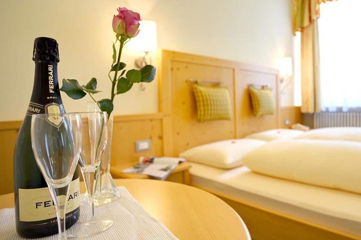 Quarto de hotel com cama, mesa de cabeceira com garrafa de vinho e copos, uma rosa e um abajur.