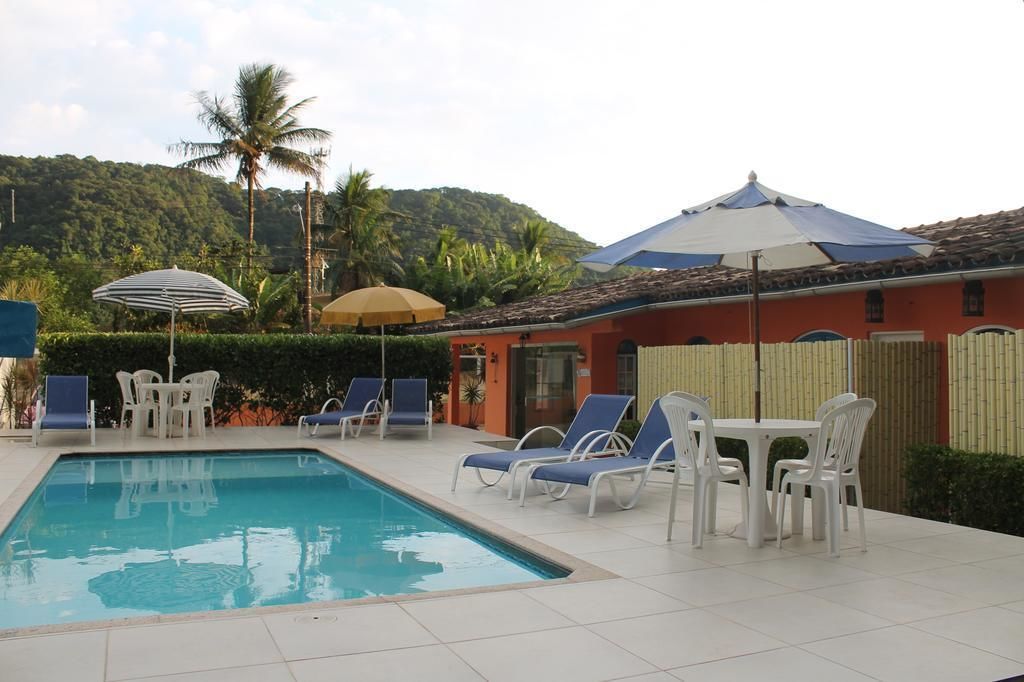 Piscina com espreguiçadeiras e guarda-sóis, cercada por um edifício com telhado de azulejos e vista para uma colina arborizada.