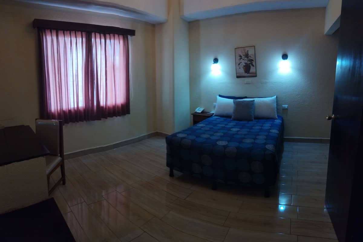 Quarto de hotel com cama, mesa de cabeceira, escrivaninha e janela com cortinas.
