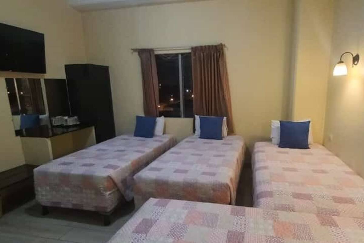 Quarto de hotel com três camas, televisão fixada na parede, escrivaninha com cadeira e janela com cortinas.