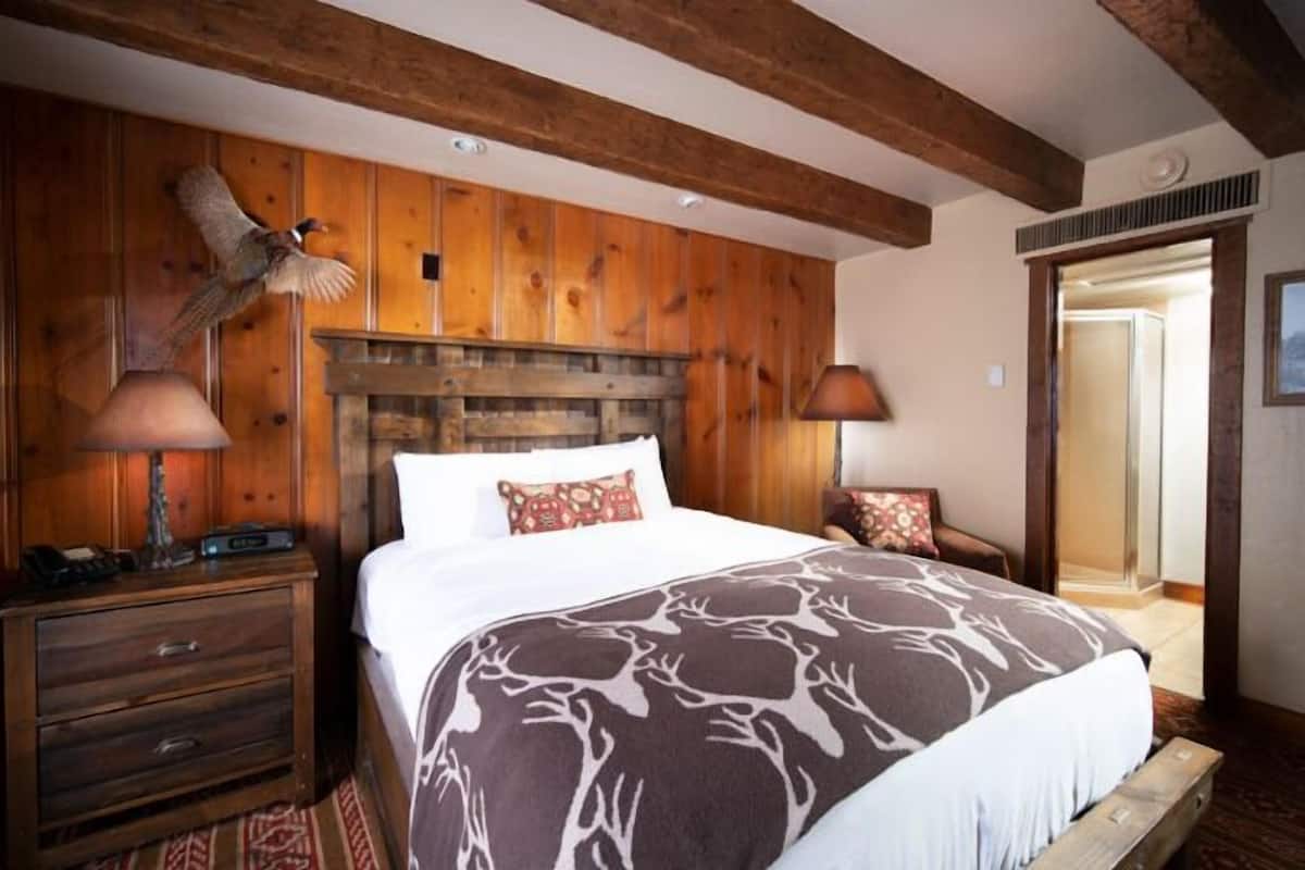 Um quarto com cabeceira de madeira, cama com colcha estampada, mesa de cabeceira com abajur e relógio fixado na parede.