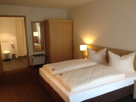 Quarto de hotel com uma cama, guarda-roupa, espelho, mesa de cabeceira com abajur e banheiro visível por uma porta aberta.