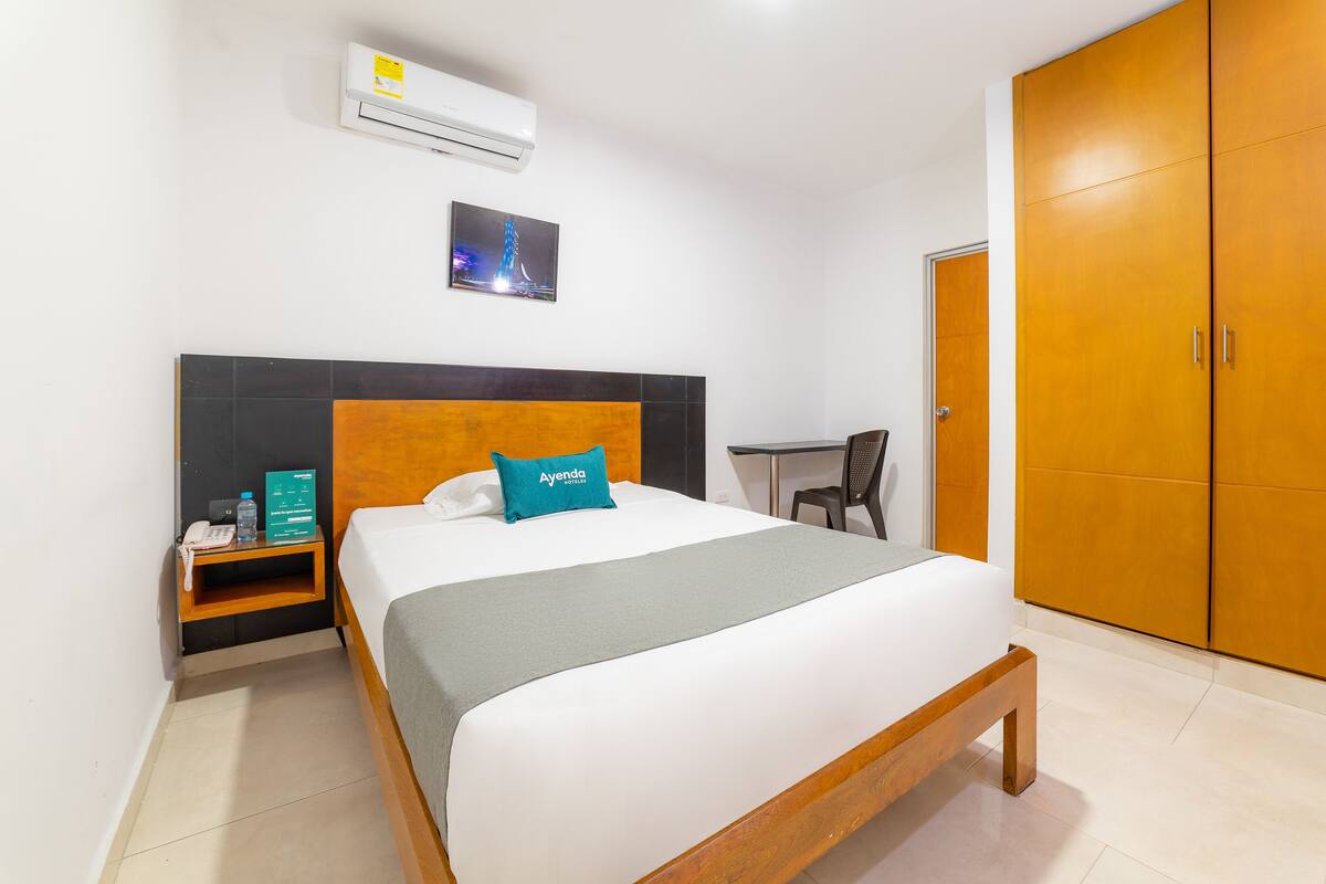 Quarto de hotel com cama, mesa de cabeceira, ar-condicionado, escrivaninha e guarda-roupa.