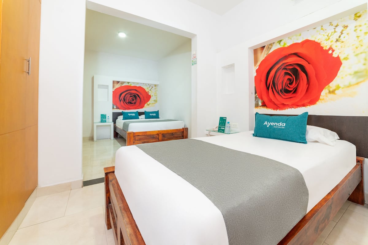 Quarto de hotel com uma cama grande, cabeceira de madeira, manta cinza e almofada com o nome do hotel.